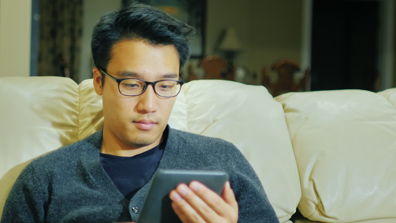 Premium stock video - A young korean man reads an e-book on the couch